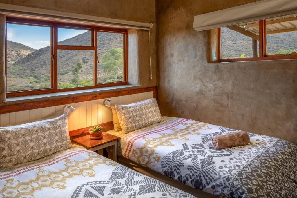 Quarto com duas camas, uma janela com vista para colinas, uma luminária e uma pequena mesa com uma planta.