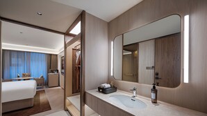 Quarto de hotel moderno com uma cama grande, banheiro com um espelho amplo e uma escrivaninha com cadeira.