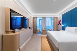 Quarto de hotel moderno com uma televisão de tela grande, uma cama, uma escrivaninha com cadeira e vista para a cidade através de amplas janelas.
