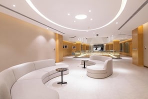 Um hall de hotel moderno com um sofá branco curvo, uma mesa redonda pequena e um corrimão de vidro ao fundo.