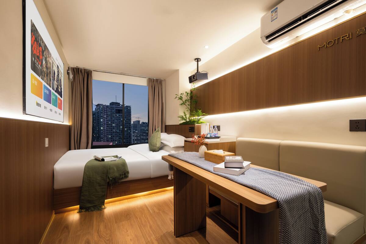 Quarto de hotel moderno com cama, escrivaninha, sofá, planta em vaso e vista para a cidade.