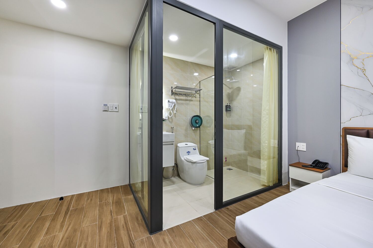 Quarto de hotel moderno com chuveiro em cabine de vidro, piso de madeira, cama com roupa de cama branca e mesa de cabeceira com telefone.