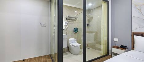 Quarto de hotel moderno com chuveiro em cabine de vidro, piso de madeira, cama com roupa de cama branca e mesa de cabeceira com telefone.