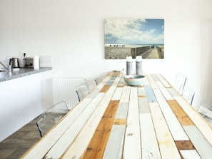 Uma área de refeições com uma mesa de madeira comprida, cadeiras, um radiador branco, uma bancada de cozinha com eletrodomésticos e um quadro com uma cena de praia na parede.