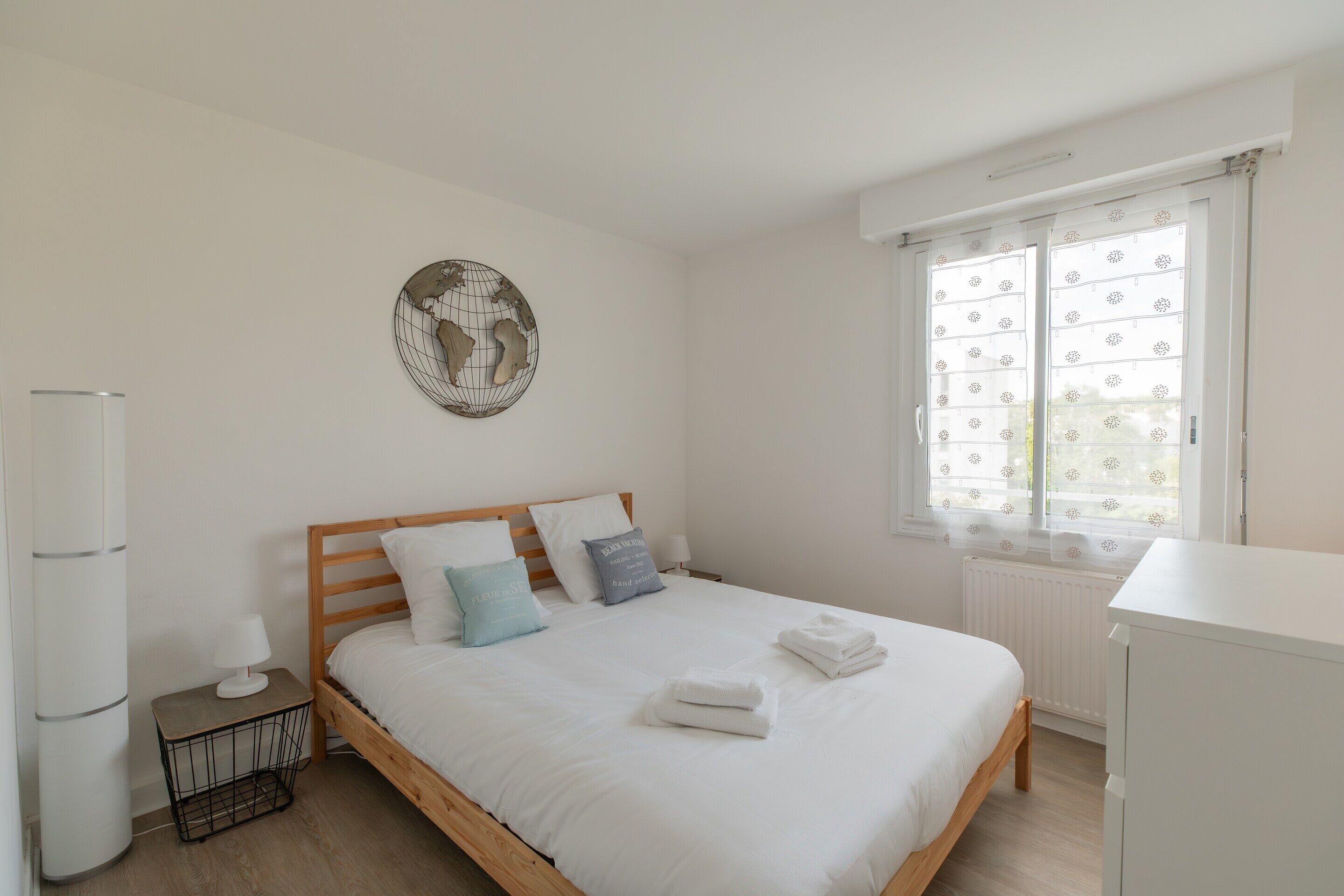 Quarto com cama de madeira, roupa de cama branca, um globo decorativo, radiador e janela com cortinas estampadas.