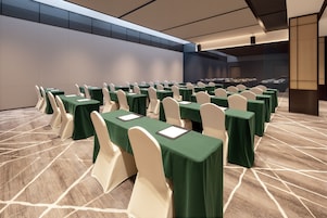 Sala de conferência com fileiras de mesas e cadeiras, piso estampado e uma janela grande.
