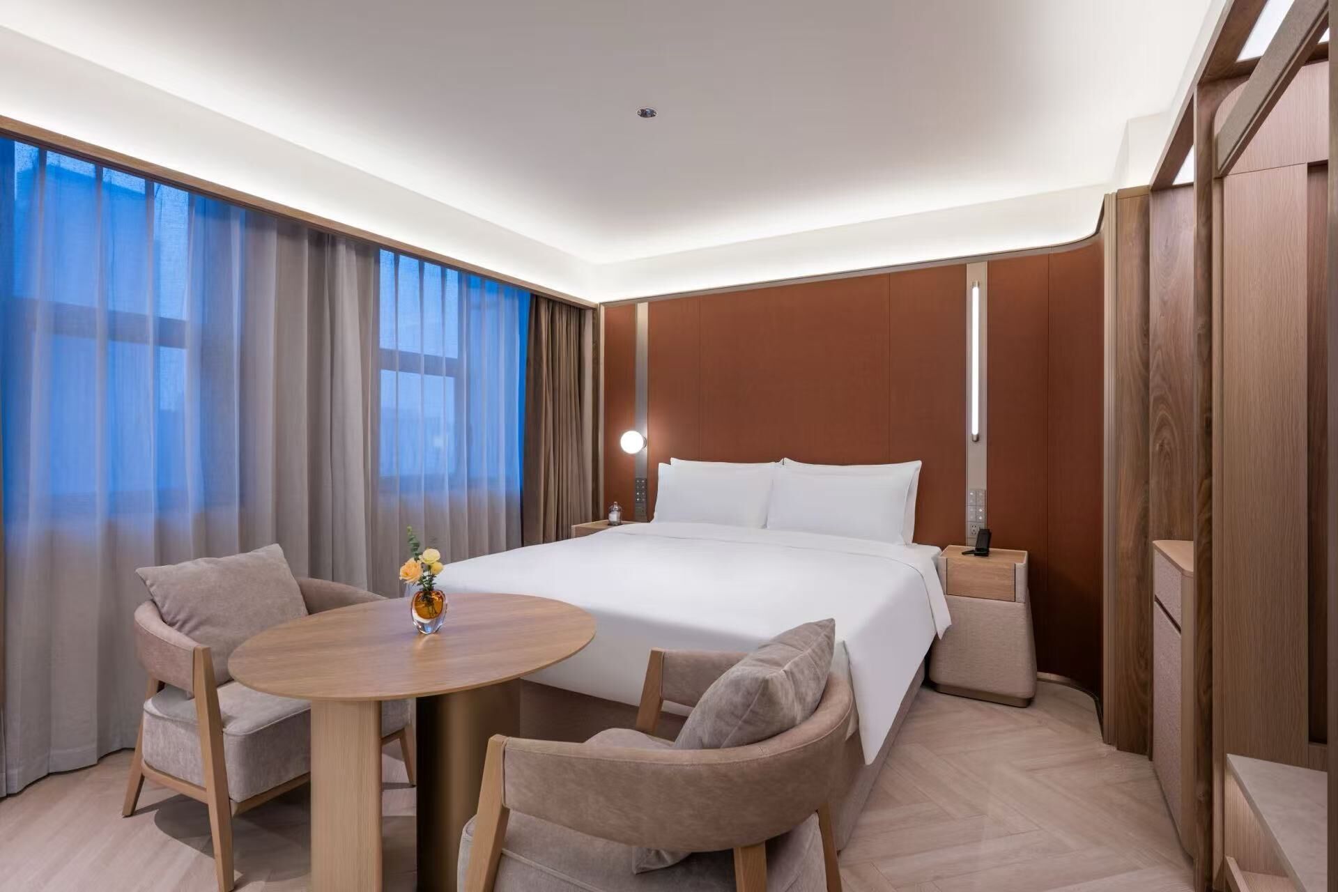 Quarto de hotel com uma cama, duas cadeiras, uma mesinha, um criado-mudo e uma janela grande com cortinas.
