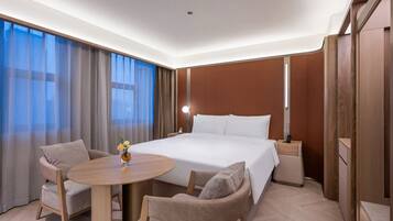 Quarto de hotel com uma cama, duas cadeiras, uma mesinha, um criado-mudo e uma janela grande com cortinas.