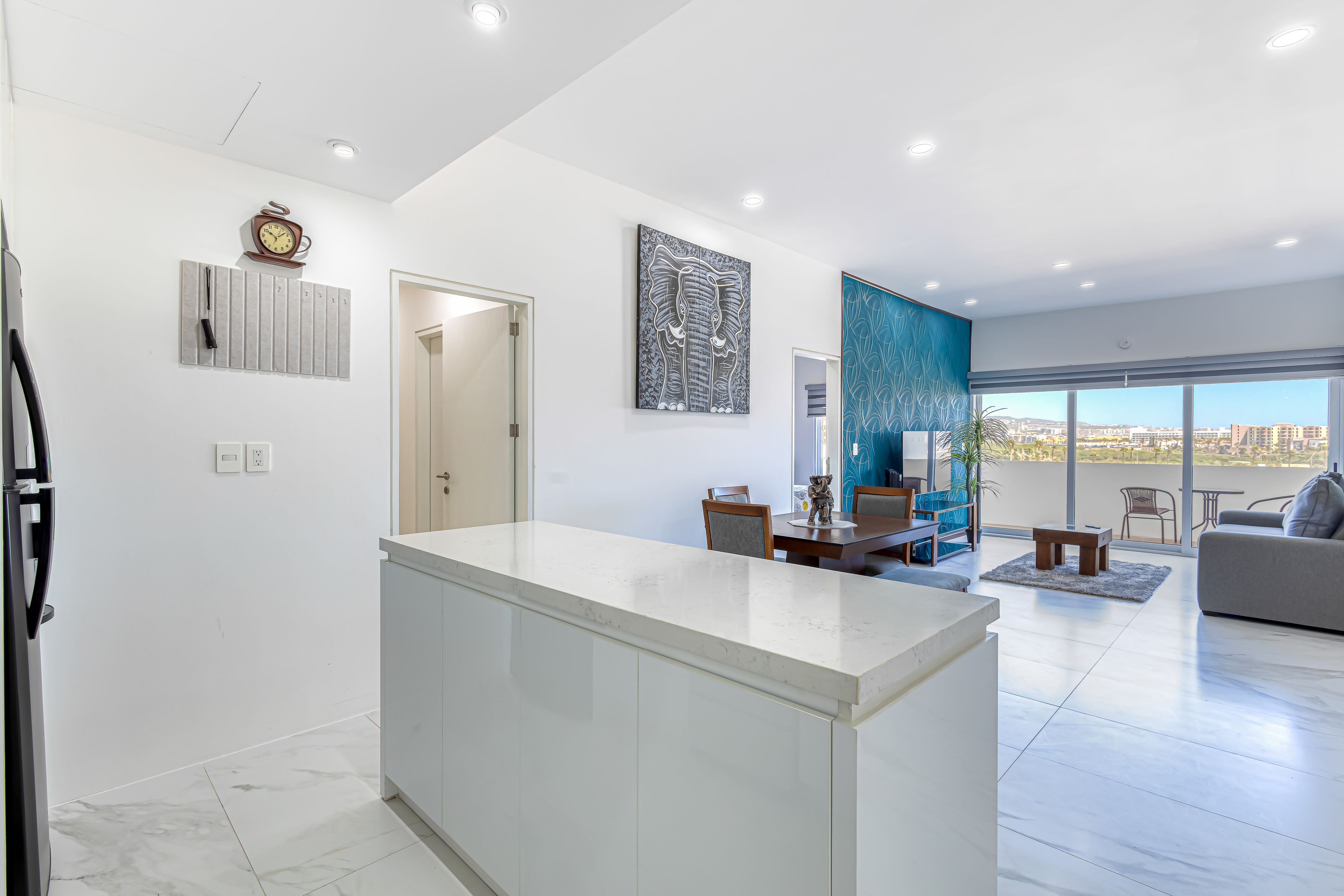 Cozinha moderna com ilha central, área de jantar com mesa e cadeiras, e sala de estar com sofá e vista da cidade.