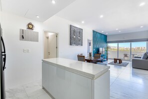 Cozinha moderna com ilha central, área de jantar com mesa e cadeiras, e sala de estar com sofá e vista da cidade.
