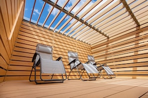 Um deck de madeira banhado pelo sol, com três cadeiras de descanso e um céu azul límpido.