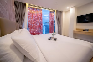 Quarto de hotel com cama grande, televisão de tela plana, mesa de cabeceira e janela com cortinas.