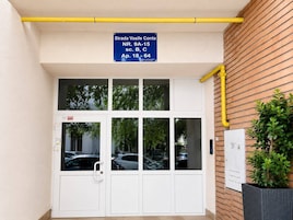 Uma entrada com portas duplas, uma placa acima, um cano amarelo e um vaso de planta ao lado da porta.