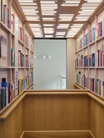 Interior de biblioteca com estantes em ambos os lados, um corredor central e uma escadaria de madeira.