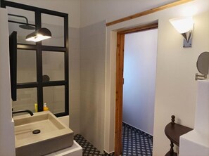 Casa de banho com lavatório, espelho, chuveiro e porta que dá para outro espaço.