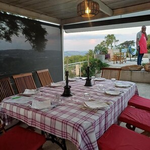 Uma área de refeições ao ar livre com uma mesa posta para quatro pessoas, rodeada por cadeiras e bancos, com vista para a paisagem.