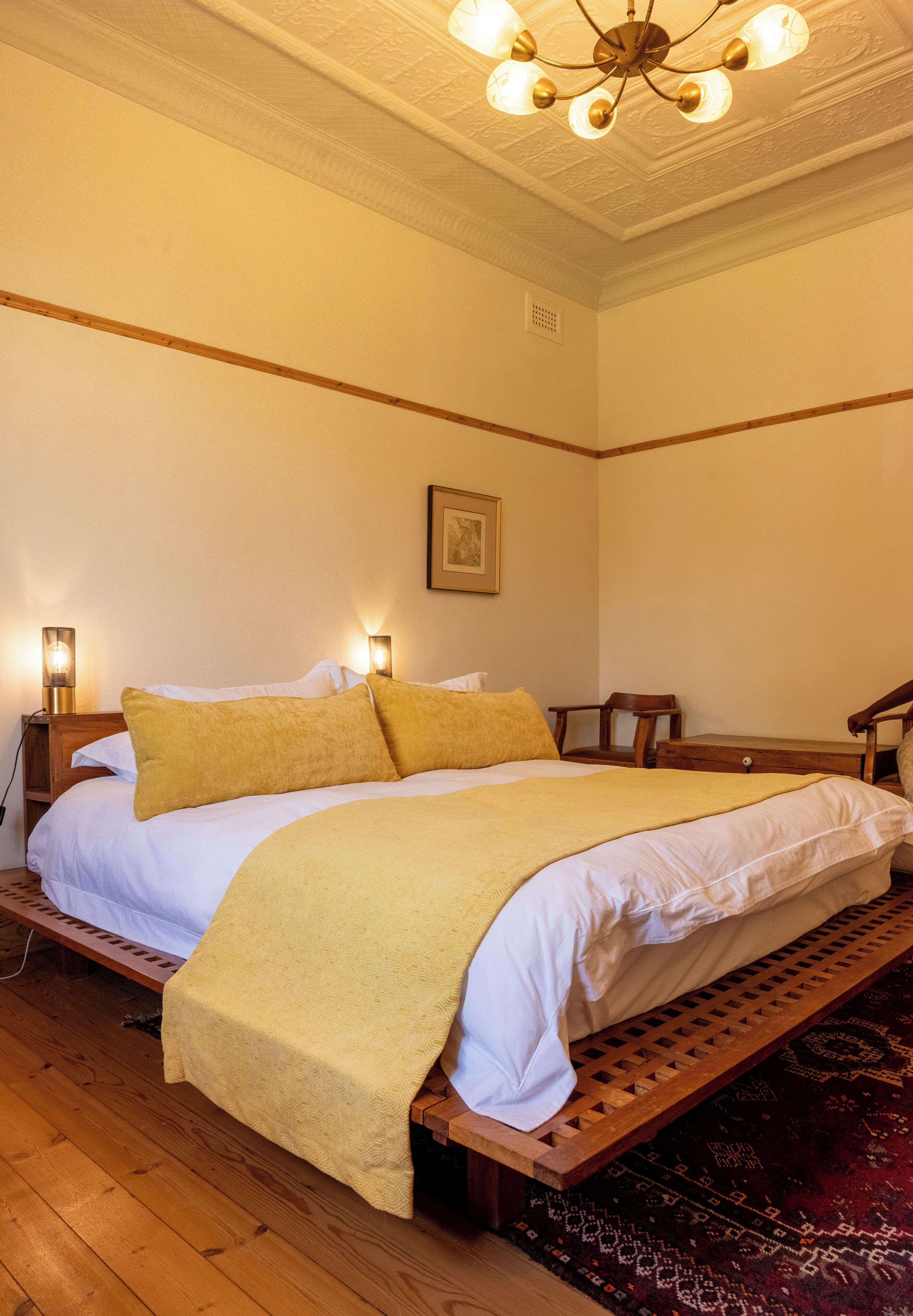 Quarto com cama de madeira, roupa de cama branca, manta amarela, luminária no teto e tapete estampado.
