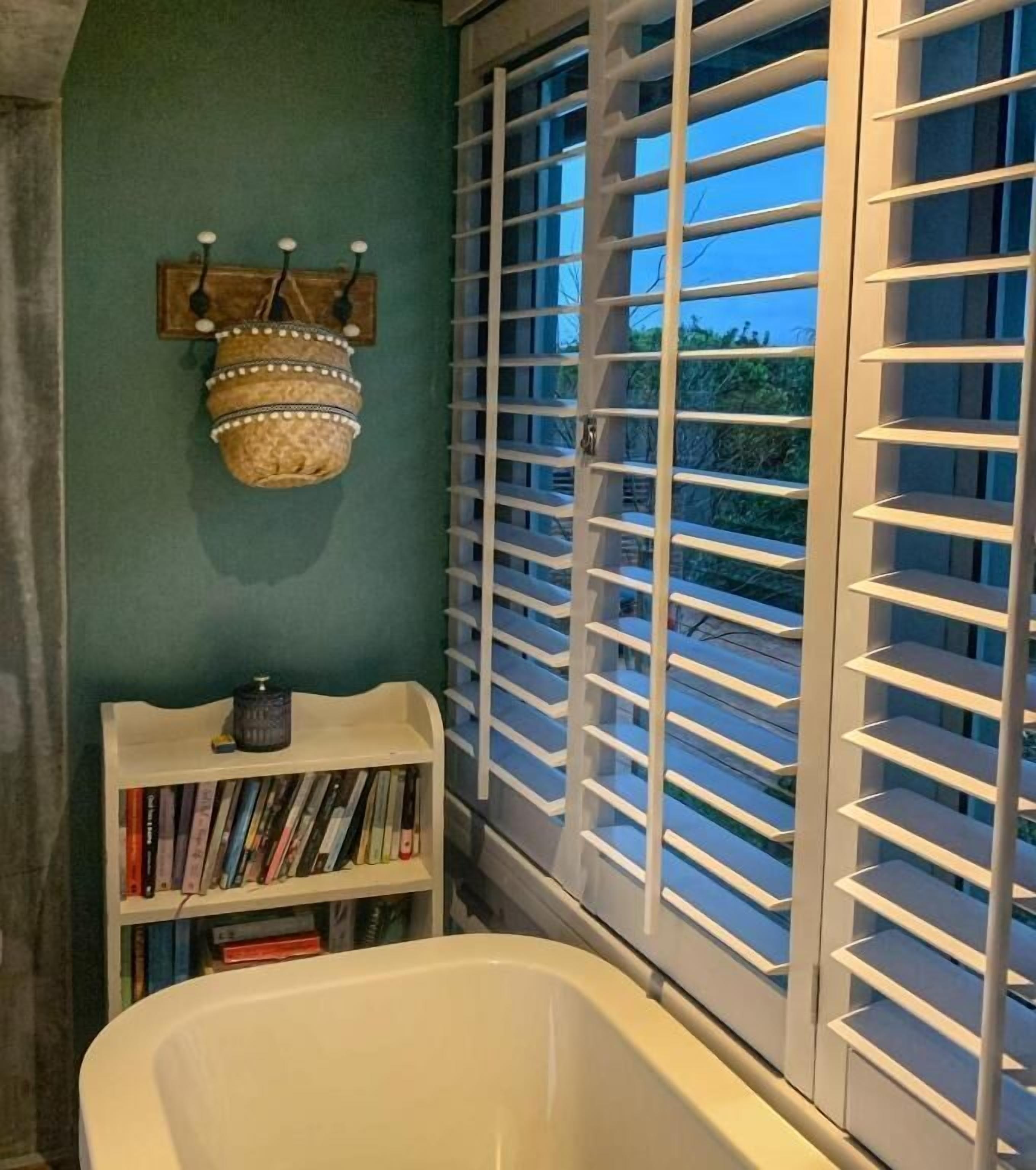 Casa de banho com banheira, prateleira com livros, cesto decorativo e janela com persianas.