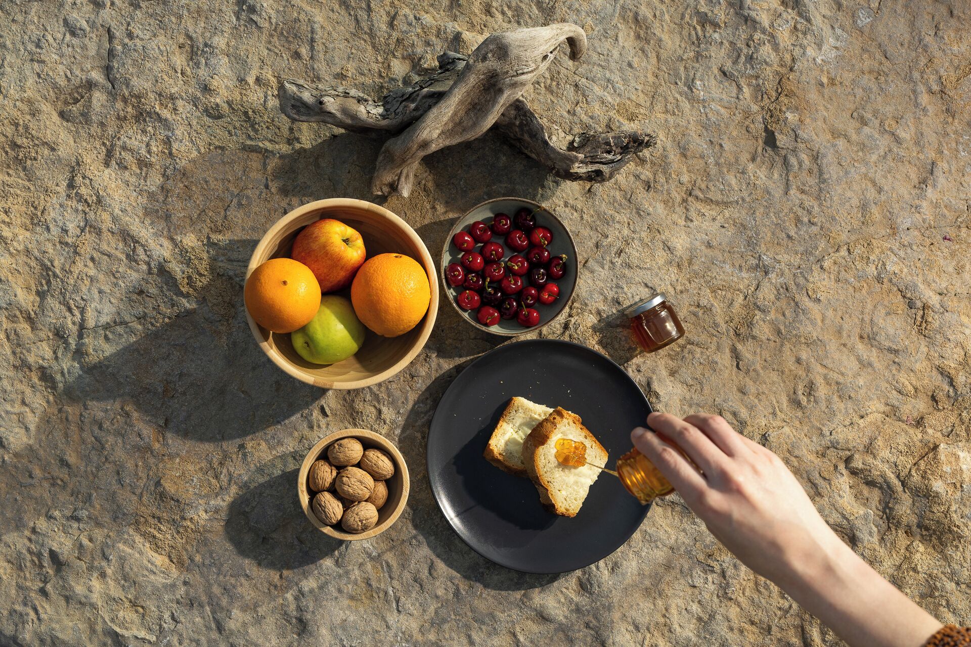 Uma tigela de madeira com frutas, um prato com torradas e um pote de mel sobre uma superfície de pedra.