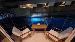 Varanda com divisória de vidro, duas cadeiras brancas acolchoadas, uma mesa de madeira e um fundo iluminado em azul.