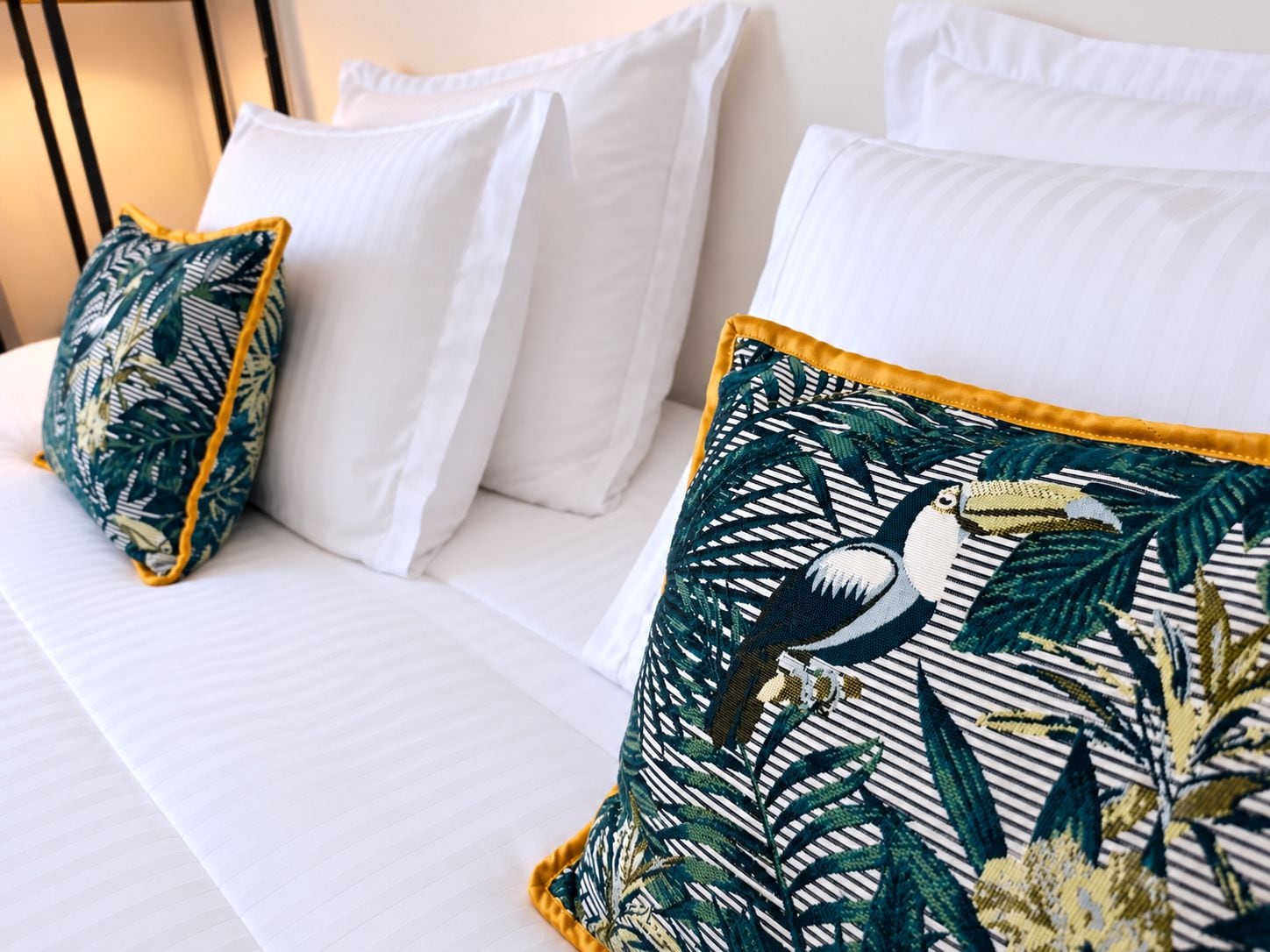 Cama com lençóis brancos e duas almofadas decorativas com estampas de folhas tropicais.