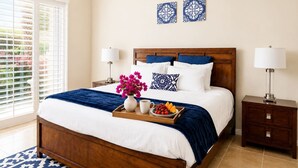 Quarto com cama de madeira, roupa de cama branca, manta azul decorativa, bandeja com frutas e flores, duas luminárias de cabeceira e uma janela com persianas.