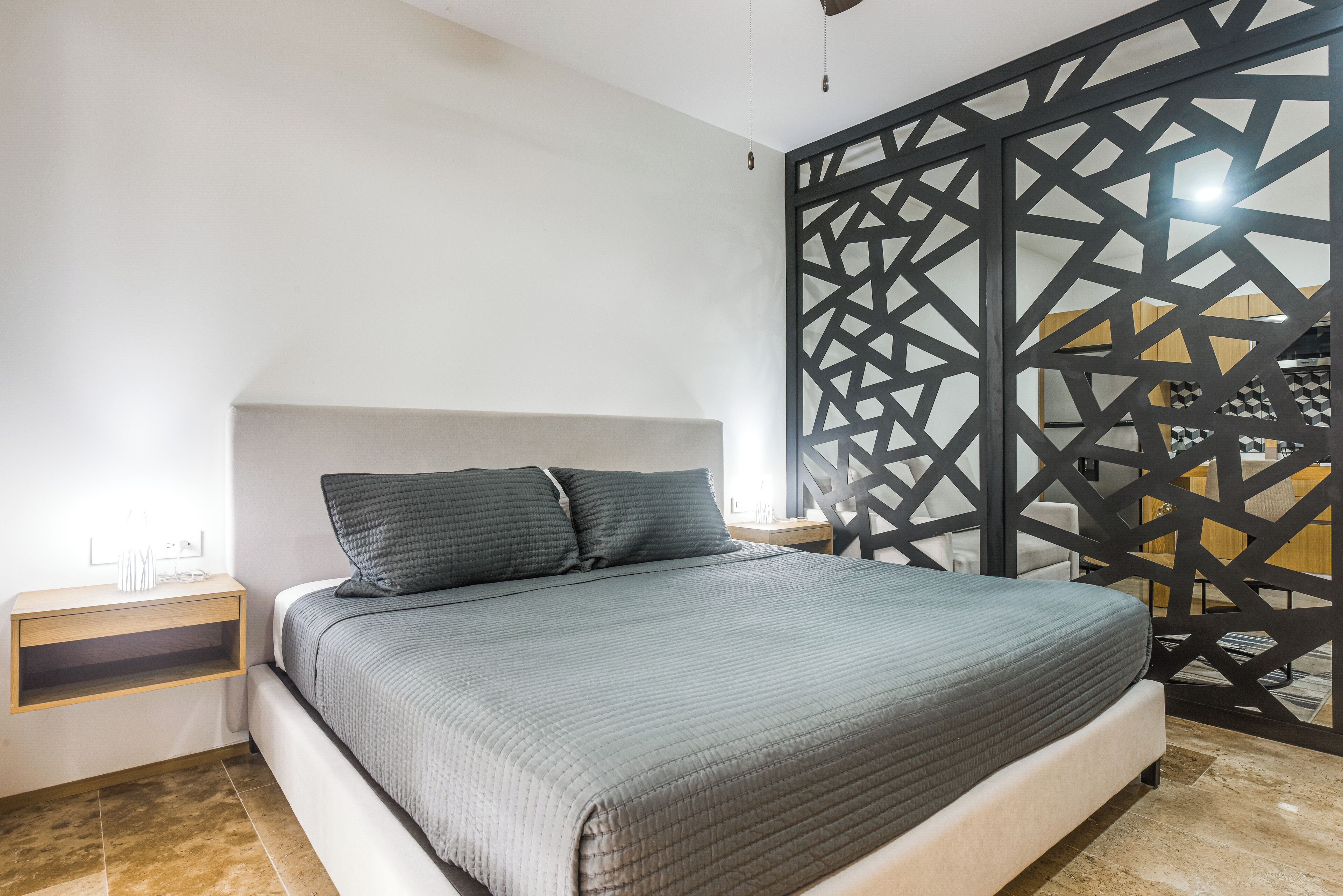 Quarto moderno com cama, mesa de cabeceira, prateleira na parede e divisória com padrão geométrico.