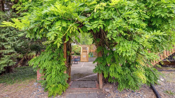 Um arco de madeira dá acesso a um pátio coberto, com um muro de tijolos e uma cerca de madeira ao fundo.