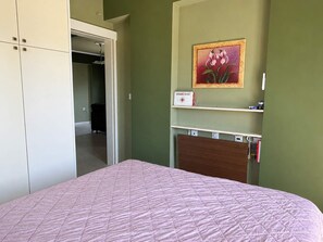 Quarto com cama, um quadro, prateleira com objetos e porta que dá para outro cômodo.