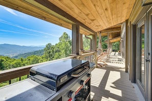 Um deck coberto ao ar livre com churrasqueira, guarda-corpo de madeira e vista para as montanhas.