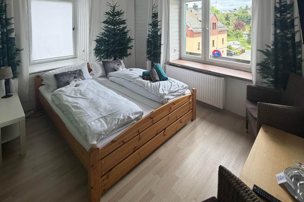 Quarto com cama de madeira, janela com vista para prédios, radiador e detector de fumaça no teto.