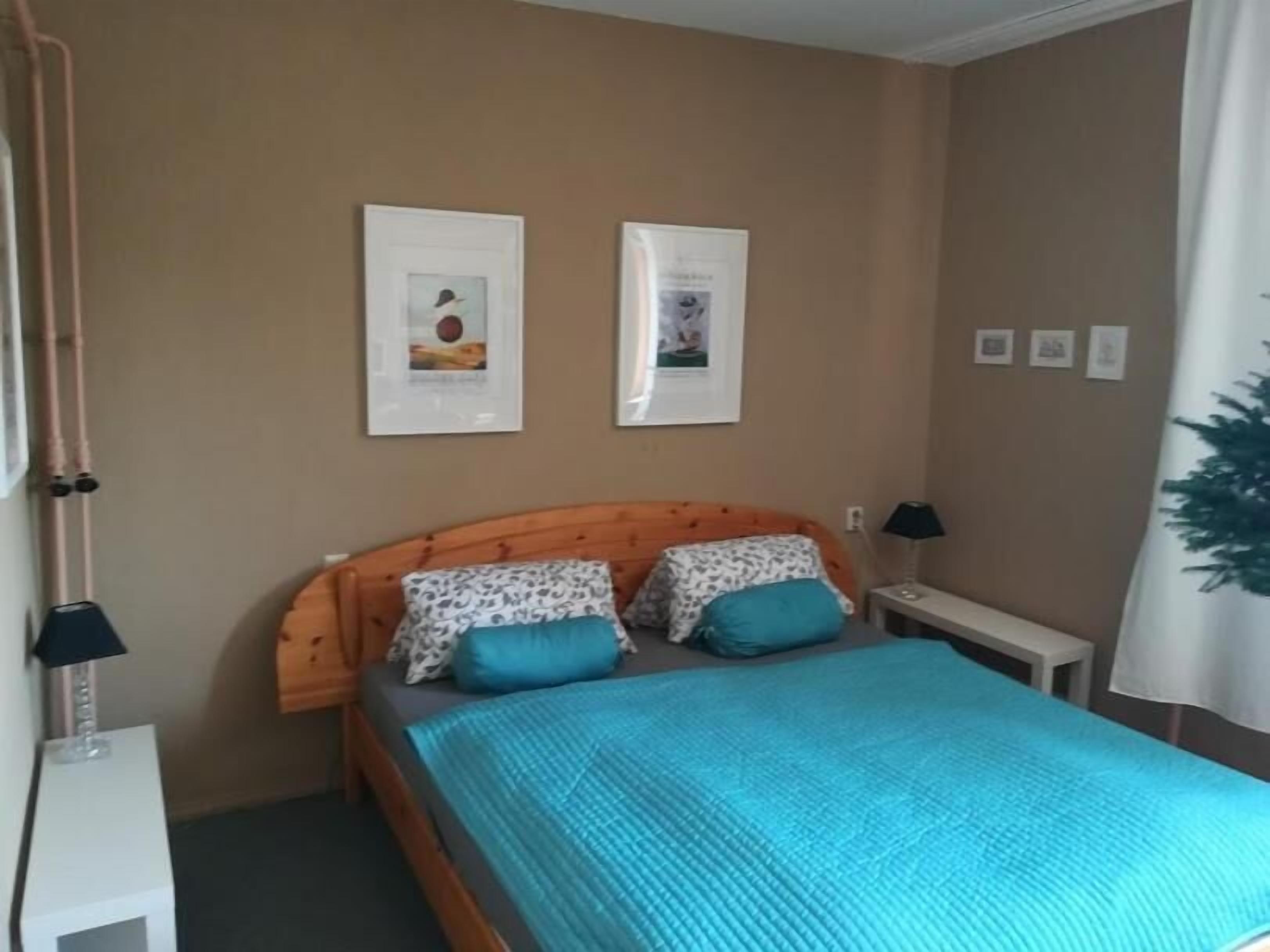 Quarto com cama, duas imagens emolduradas na parede, mesa de cabeceira com abajur e janela com cortinas.