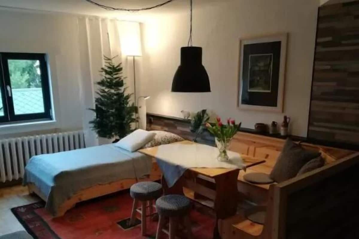 Quarto pequeno com cama, uma mesinha com banquinhos, radiador, janela e um pinheiro de Natal.