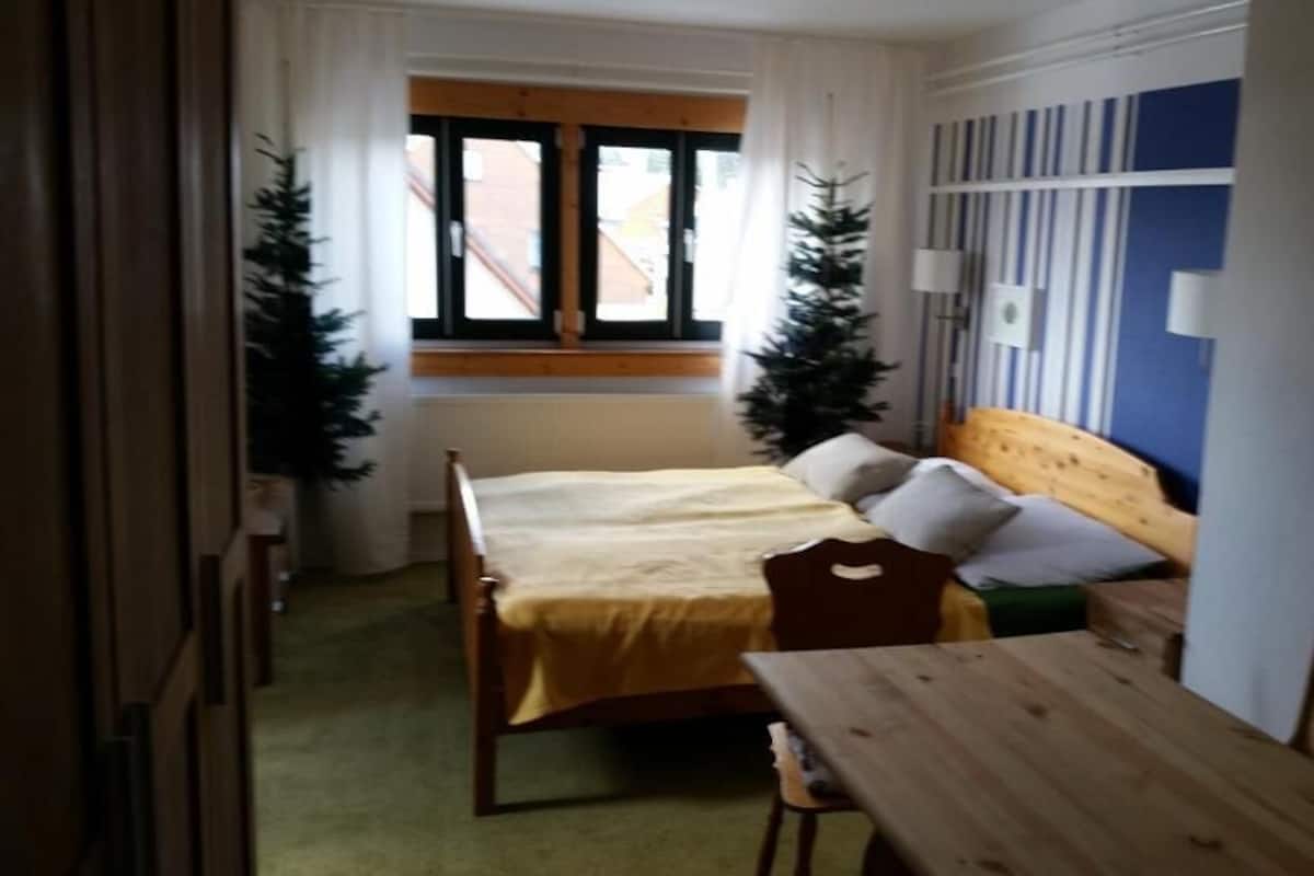 Quarto com cama, mesa de madeira, cadeira, janela com cortinas e luminária na parede.