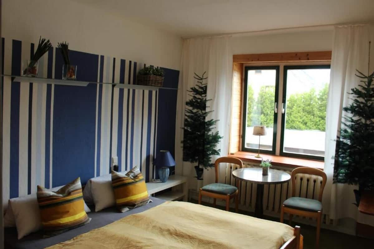 Um quarto com uma cama, duas cadeiras, uma mesa pequena, uma janela com cortinas e uma parede listrada.