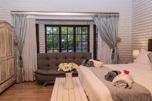 Quarto de hotel com cama grande, sofá, piso de madeira, janela com cortinas e um vaso de flores.