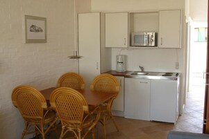 Uma cozinha compacta com armários brancos, micro-ondas, máquina de café e pia. Há uma mesa de jantar com quatro cadeiras de vime.