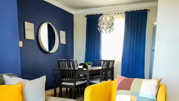 Uma sala de estar com sofá, uma poltrona amarela, uma mesa de centro com um livro, uma mesa de jantar com cadeiras e um lustre.