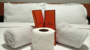 Cama bem arrumada com lençóis brancos, duas toalhas enroladas e dois kits de cortesia do hotel.