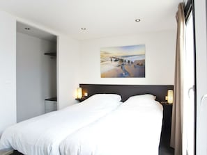 Quarto de hotel com uma cama, duas luminárias de cabeceira, um quadro com uma paisagem de praia e uma porta para o banheiro.