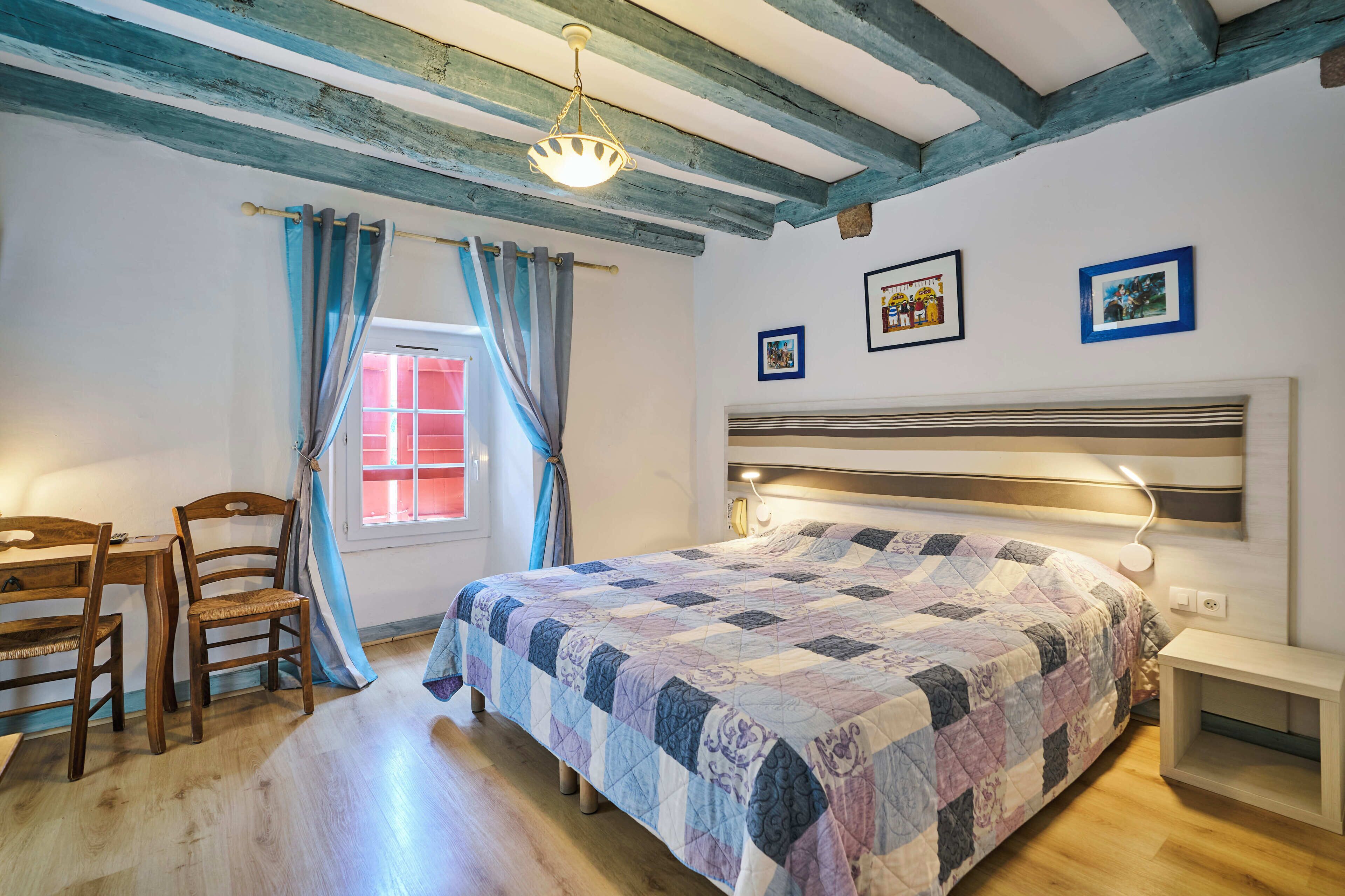 Quarto com cama, escrivaninha, cadeiras, janela com cortinas e teto com vigas aparentes.