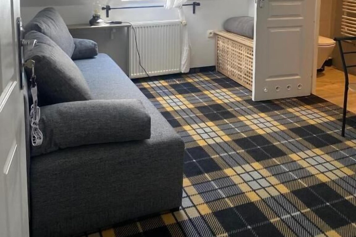 Quarto pequeno e aconchegante, com um sofá cinza, um radiador, uma cesta de roupa e um chão estampado.