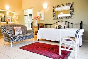 Quarto com sofá xadrez, cama com colcha branca, tapete vermelho e espelho com moldura ornamentada.