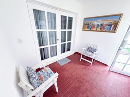 Quarto com porta branca, cadeira branca com almofada estampada, piso de azulejos vermelhos e um quadro na parede.