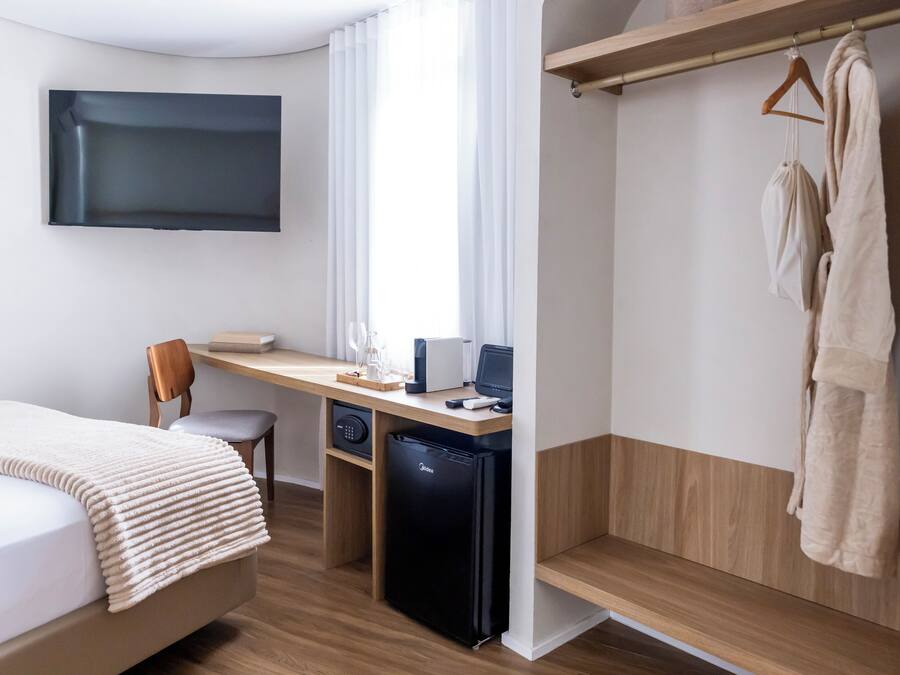 Quarto de hotel com cama, escrivaninha, cadeira, televisão e guarda-roupa.