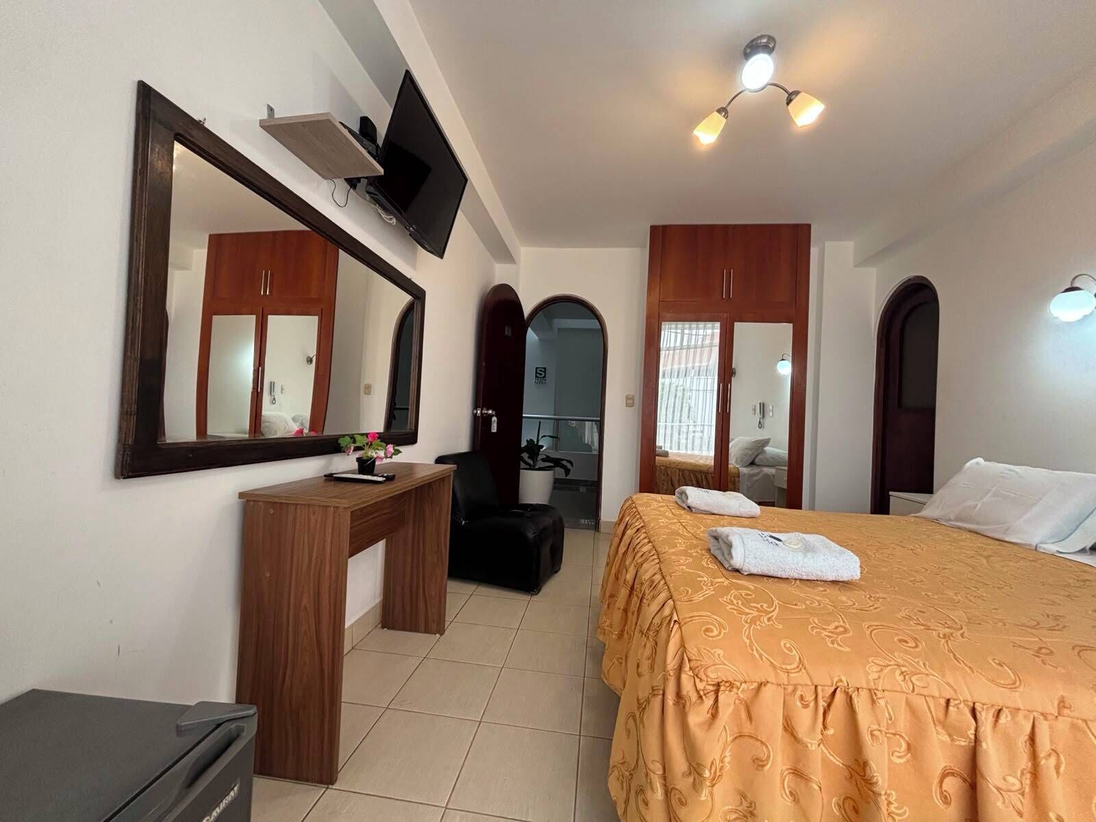 Quarto de hotel com cama, escrivaninha, cadeira, televisão e espelho.