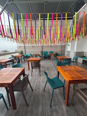 Uma área externa com mesas de madeira e cadeiras verdes, decorada com fitas coloridas.