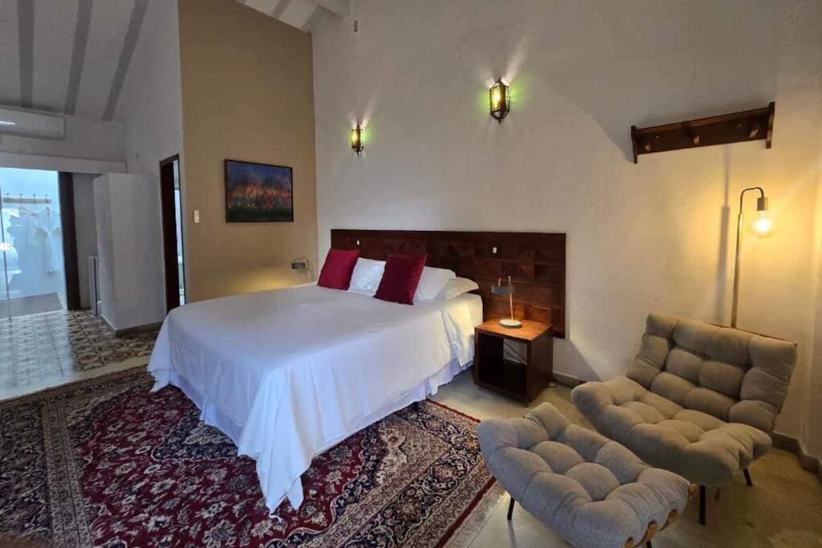 Quarto com cama grande, uma cadeira, mesa de cabeceira e luminária na parede.