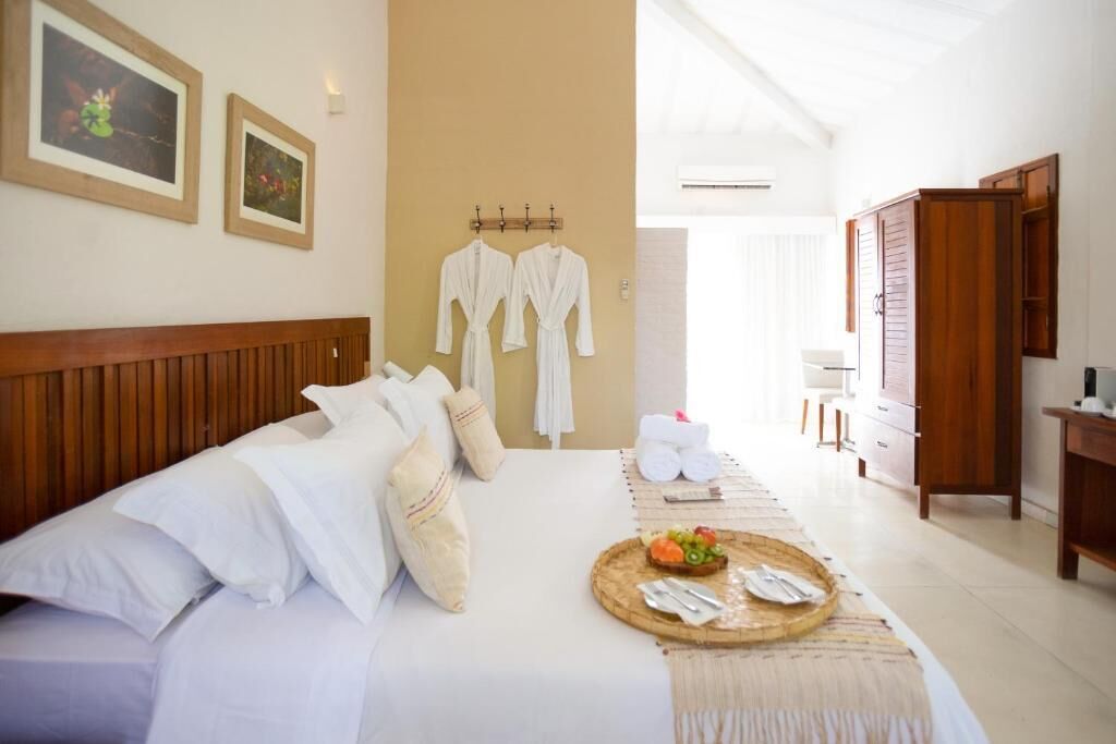 Quarto de hotel com cama, cabeceira de madeira, cabideiro fixado na parede, bandeja de café da manhã com frutas e guarda-roupa de madeira.