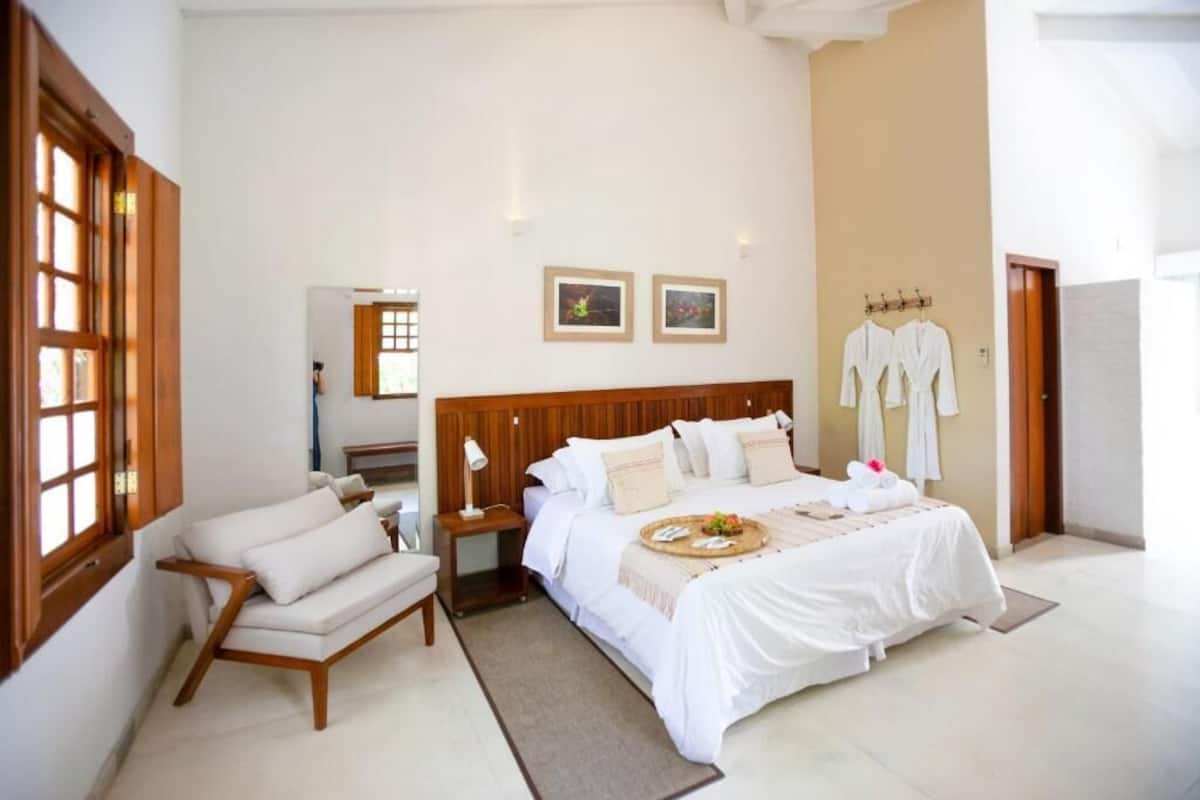 Quarto de hotel com cama, sofá, mesa de cabeceira e cabideiro fixado na parede.